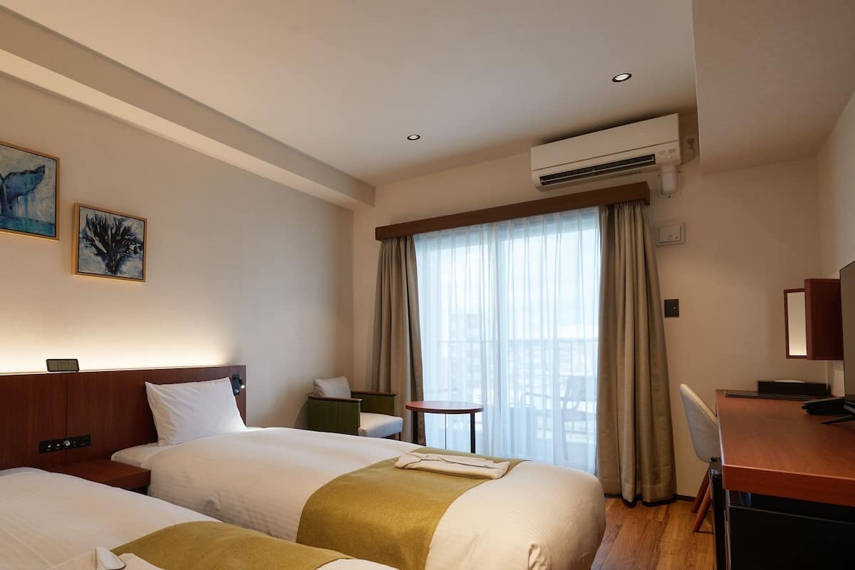 Quarto de hotel com duas camas, uma escrivaninha, uma cadeira, uma mesa pequena e uma janela com cortinas.
