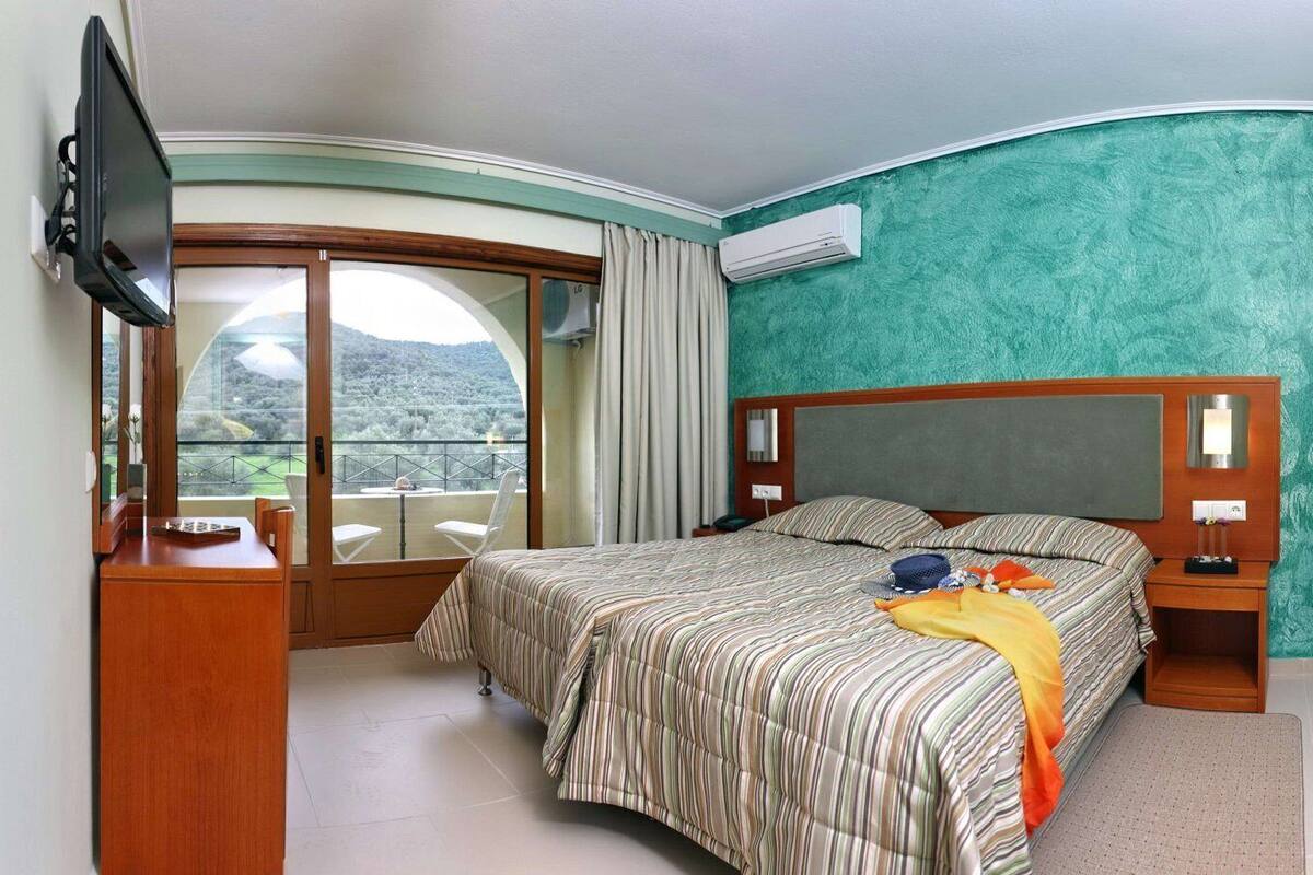 Quarto de hotel com uma cama grande, escrivaninha, cadeira, televisão e varanda com vista para as colinas.