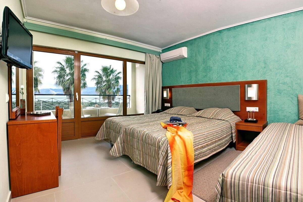 Quarto de hotel com duas camas, uma escrivaninha, uma televisão e uma varanda com vista para palmeiras e montanhas.