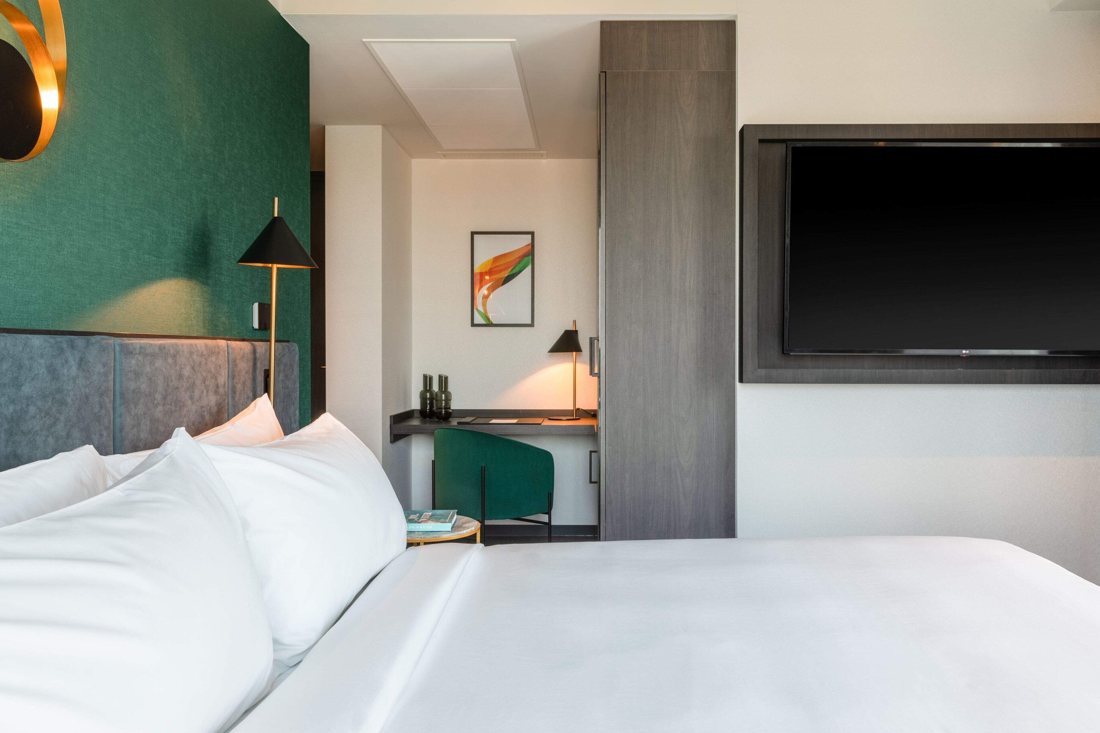 Quarto de hotel com uma cama grande, uma mesa, uma TV e uma parede verde de destaque.