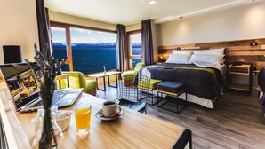Quarto de hotel com uma cama, uma escrivaninha com um laptop, uma cadeira, uma mesa com um copo de suco e vista para o mar.