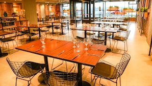 Um restaurante amplo, com várias mesas e cadeiras, grandes janelas e uma parede de tijolos.