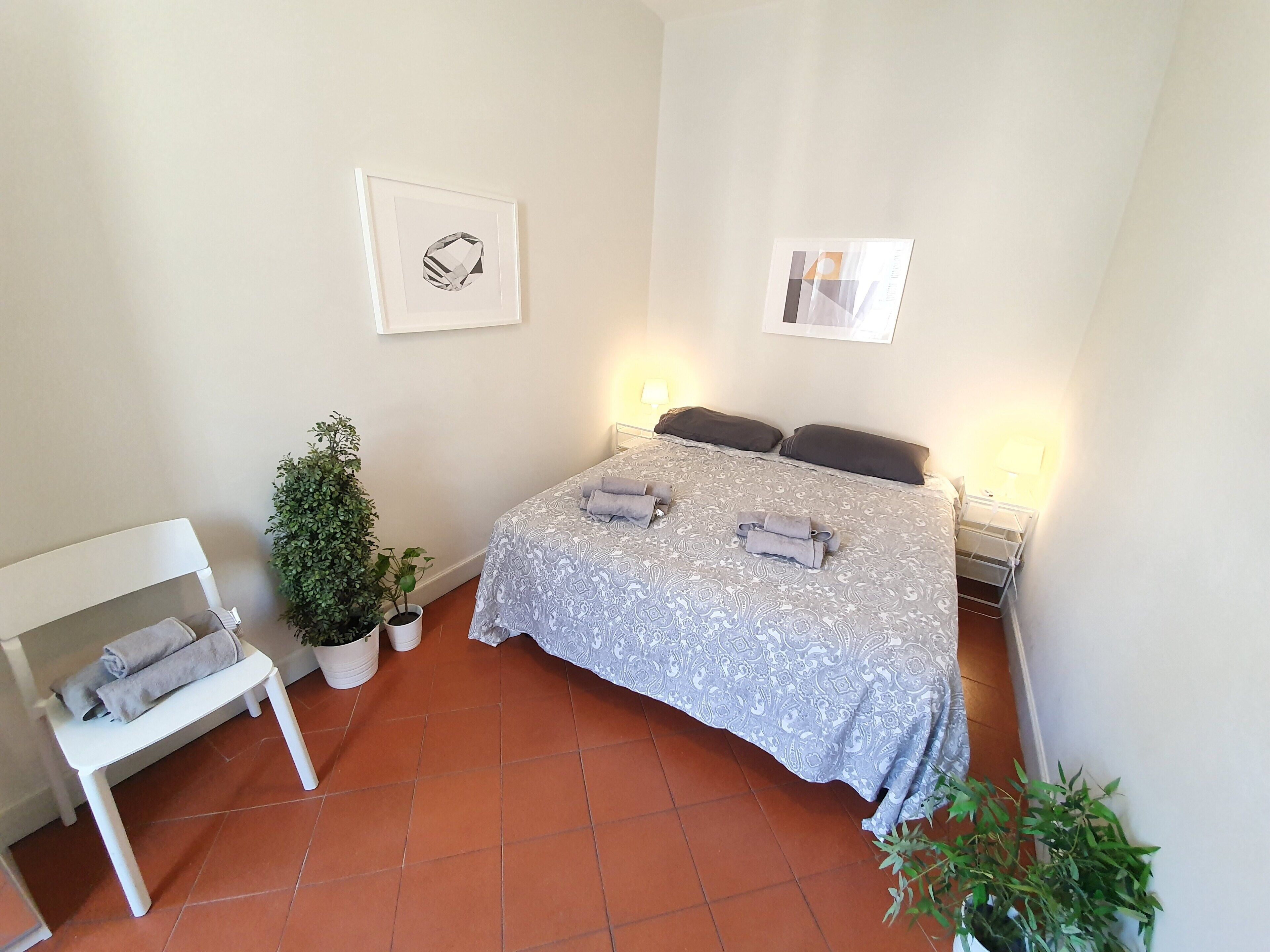 Quarto bem arrumado com cama, mesas de cabeceira, um banco e duas plantas em vasos.
