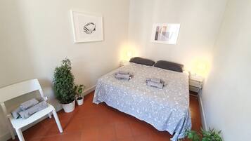 Quarto bem arrumado com cama, mesas de cabeceira, um banco e duas plantas em vasos.