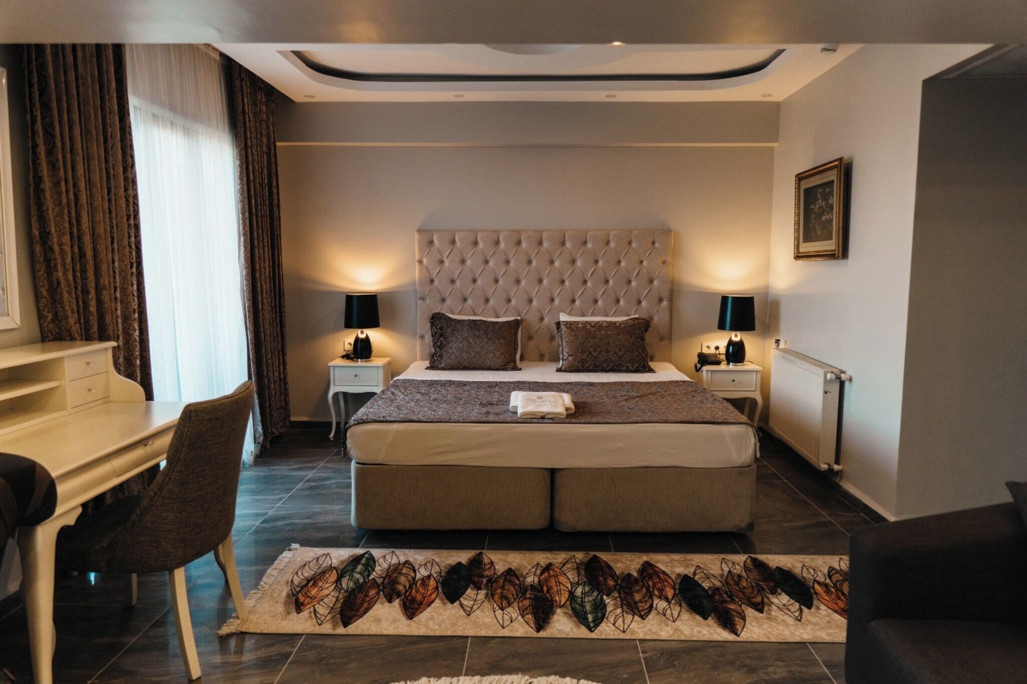 Quarto com cabeceira estofada, escrivaninha, cadeira, mesa de cabeceira com luminárias e radiador.