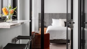 Quarto de hotel moderno com porta de vidro que dá para um quarto, uma pequena mesa com uma cadeira e um vaso com flores no peitoril da janela.