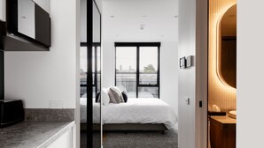Quarto moderno com uma janela grande, uma cama com travesseiros, uma mesinha de cabeceira e vista para a cidade.