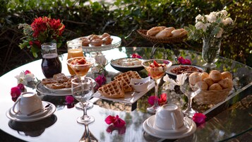 Mesa posta com diversos itens de café da manhã, incluindo waffles, pastéis e bebidas, cercada por flores e folhagem.