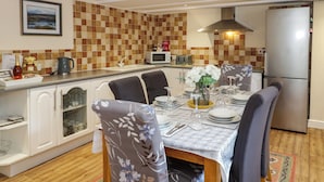 Uma sala de jantar com uma mesa posta para seis pessoas, rodeada por cadeiras com estofamento floral. Uma geladeira e um micro-ondas são visíveis ao fundo.