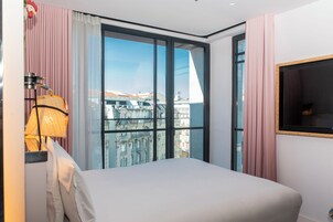 Quarto de hotel com uma janela ampla, uma cama, um abajur e uma televisão.