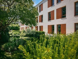 Um edifício moderno com várias janelas e um jardim bem cuidado, com diversas plantas e uma grande árvore.