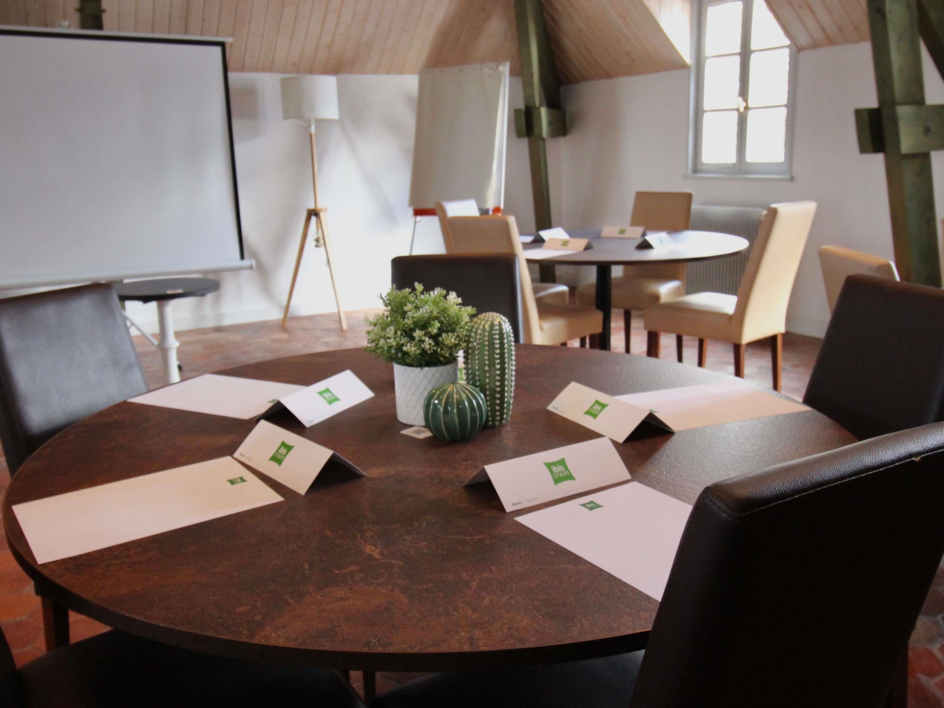 Uma sala de conferências com uma mesa redonda, cadeiras, um quadro branco e um vaso de planta.