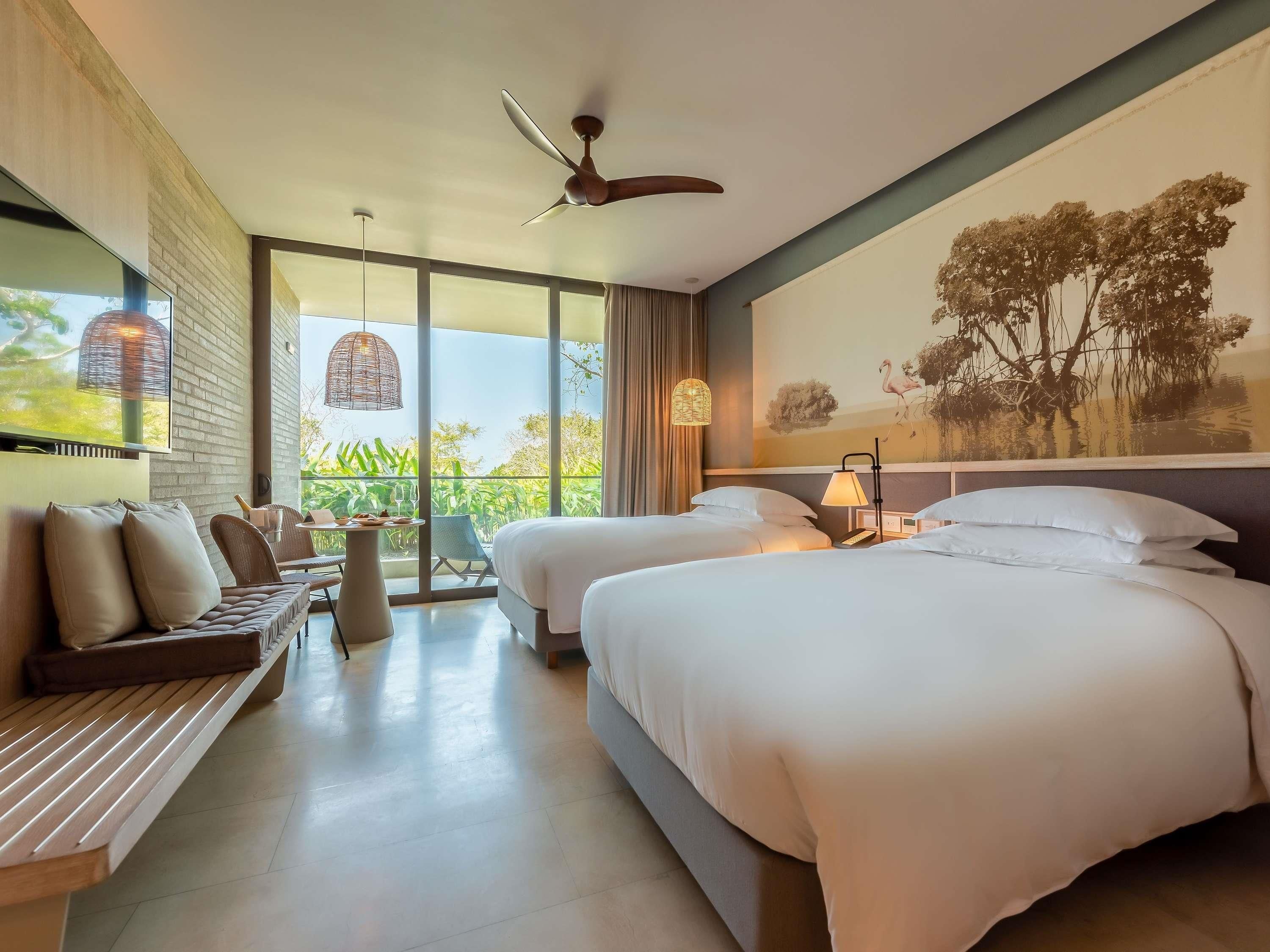 Quarto de hotel com cama, área de estar, ventoinha no teto e uma grande janela com vista para o verde.