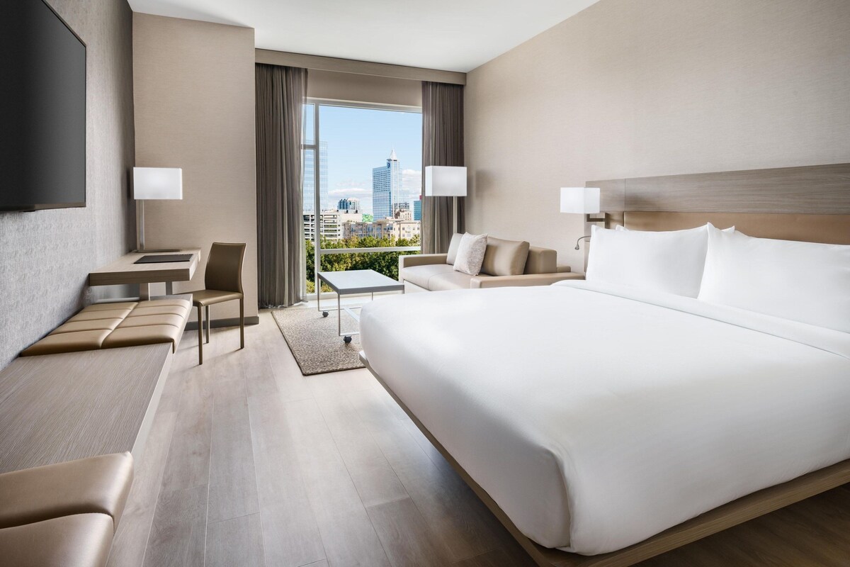 Quarto de hotel moderno com uma cama grande, uma escrivaninha, uma cadeira e um sofá. Há uma TV na parede e uma janela com vista para a cidade.