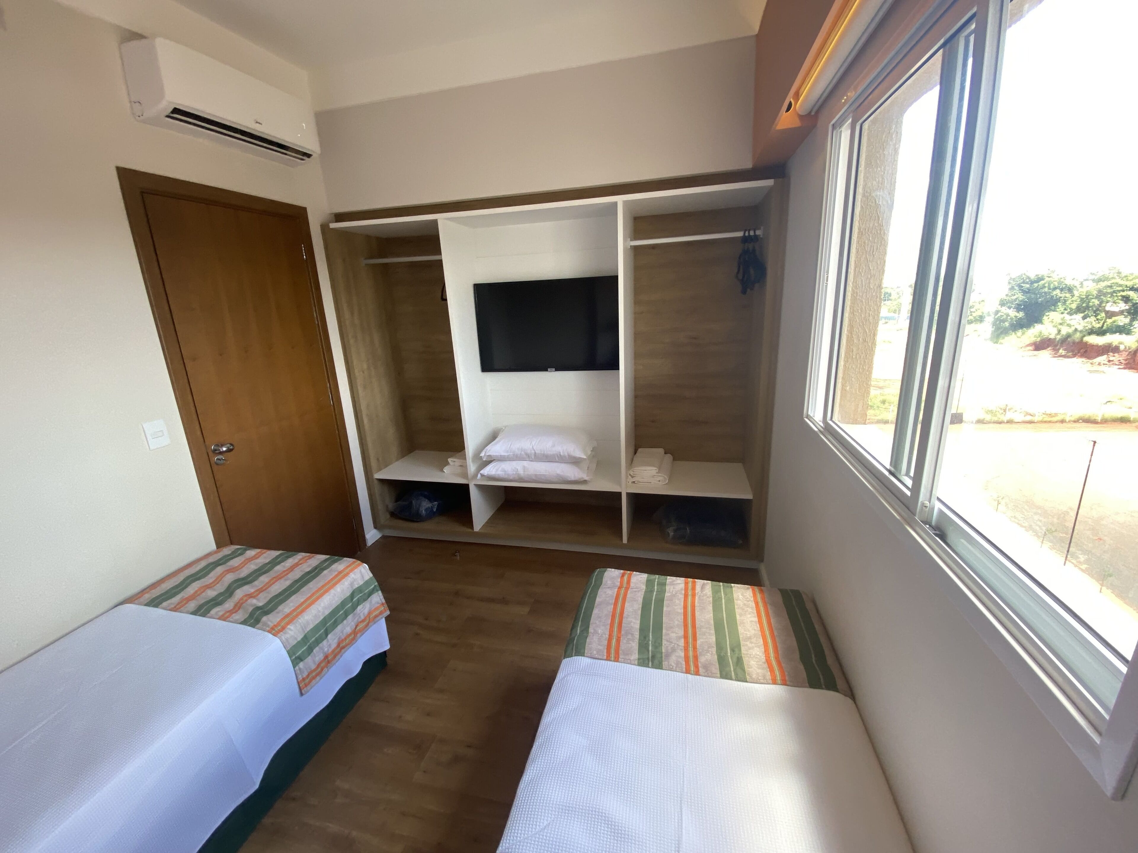 Quarto com duas camas, televisão, guarda-roupa e janela com vista para o exterior.