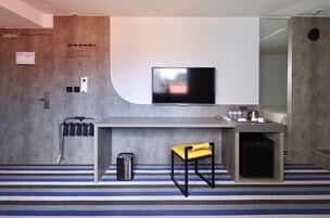 Quarto de hotel moderno com escrivaninha cinza, televisão de tela plana, uma pequena lixeira e uma cadeira amarela.