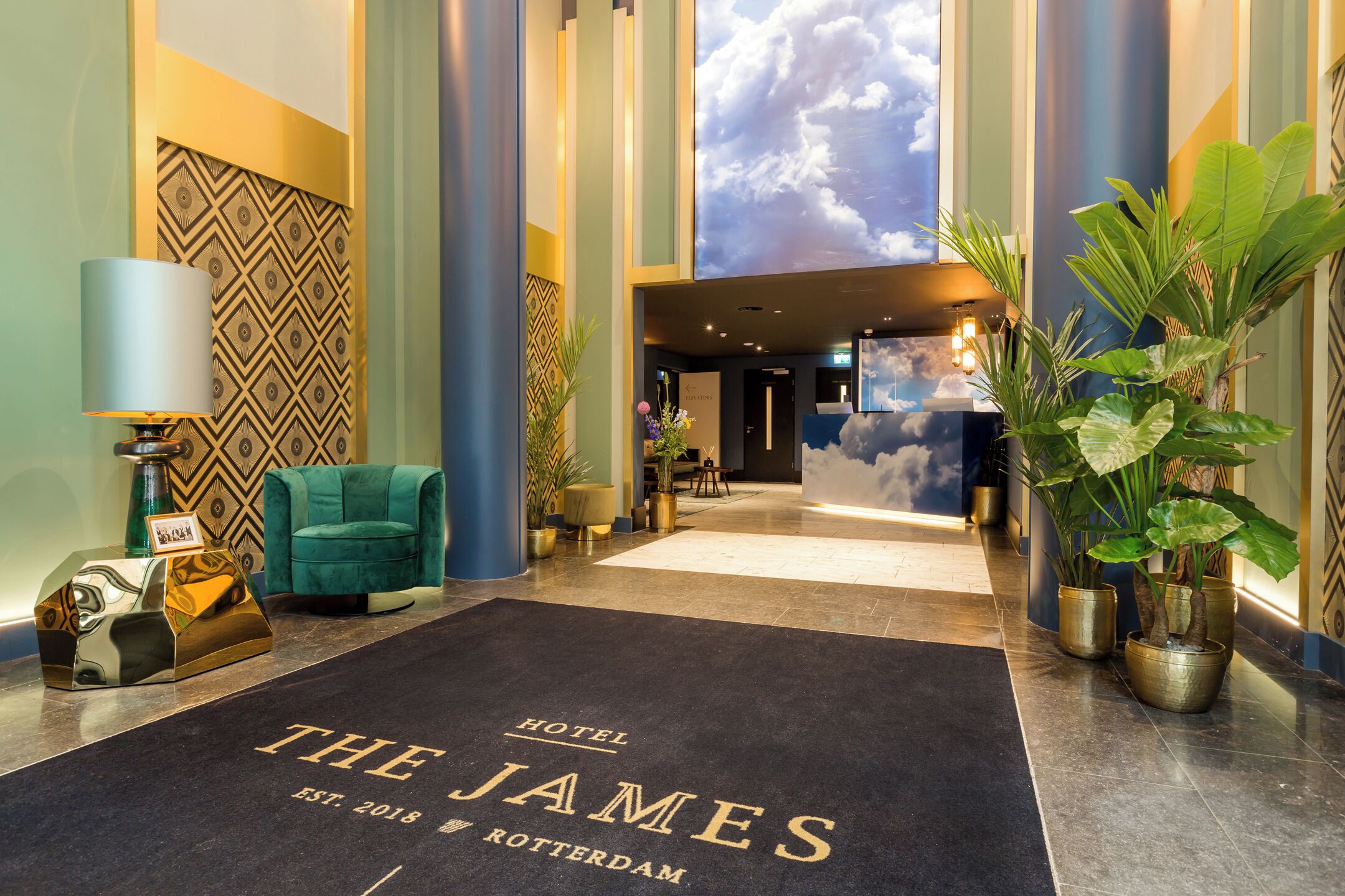 Um saguão de hotel com balcão de recepção, uma poltrona verde, plantas em vasos e um grande mural na parede.