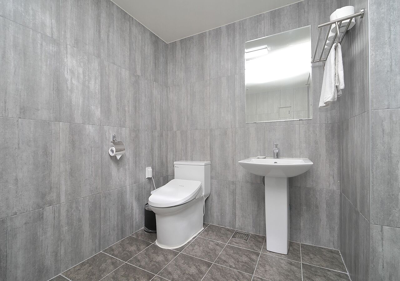 Casa de banho com sanita, lavatório e suporte para toalhas, tudo junto a uma parede revestida a azulejos cinzentos.