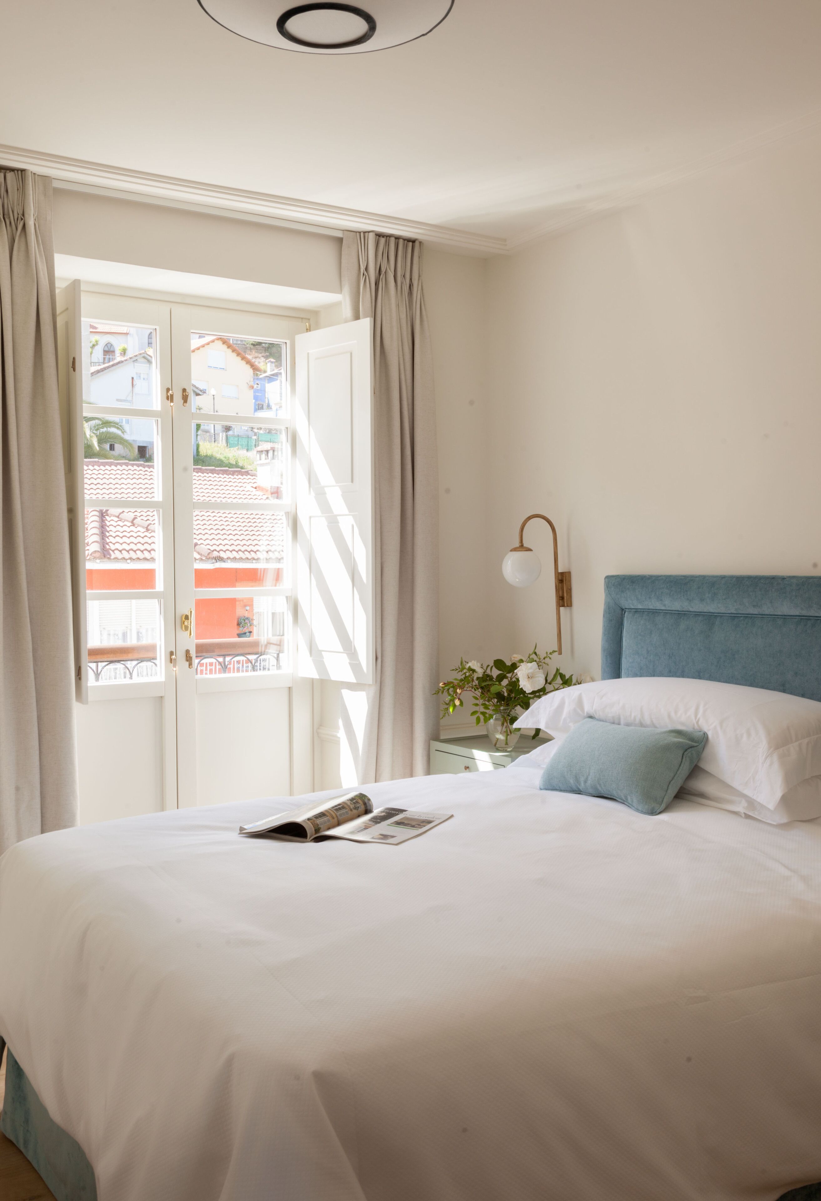 Quarto com cama, mesa de cabeceira, abajur e janela com cortinas.