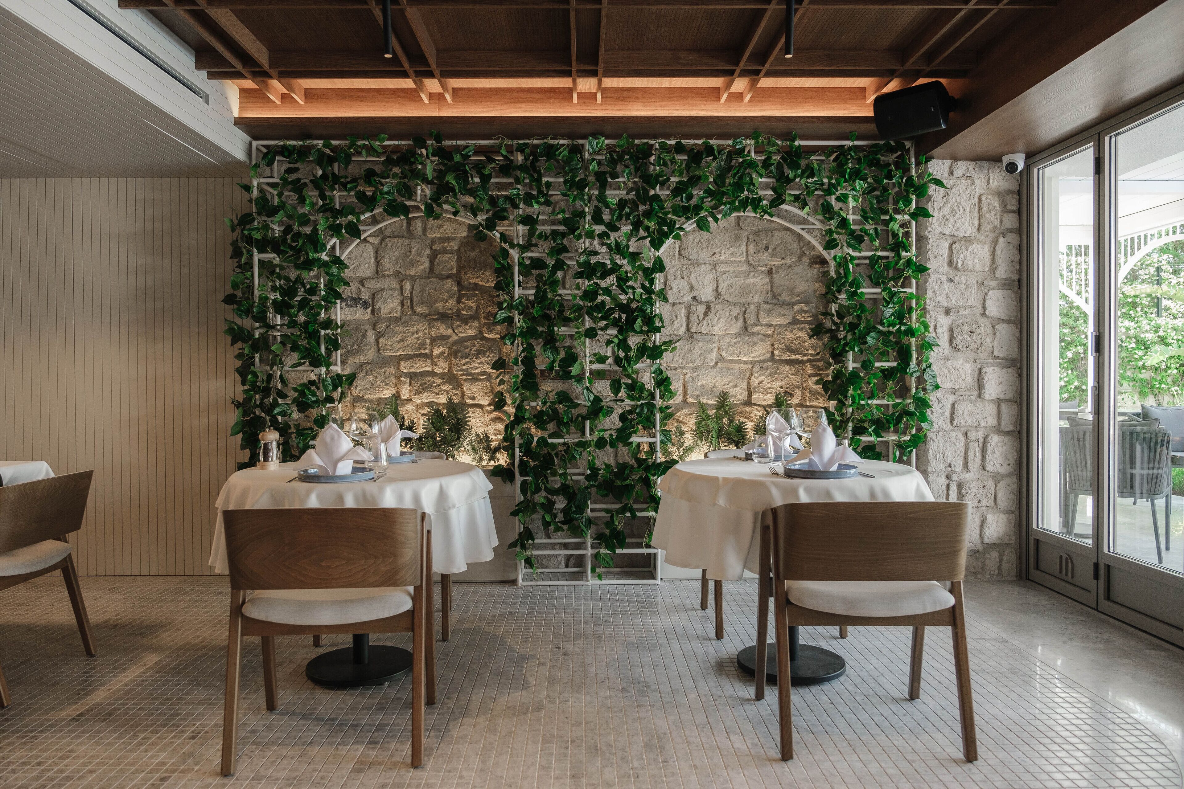 Restaurante com mesas redondas, cadeiras e uma parede de pedra com vegetação.