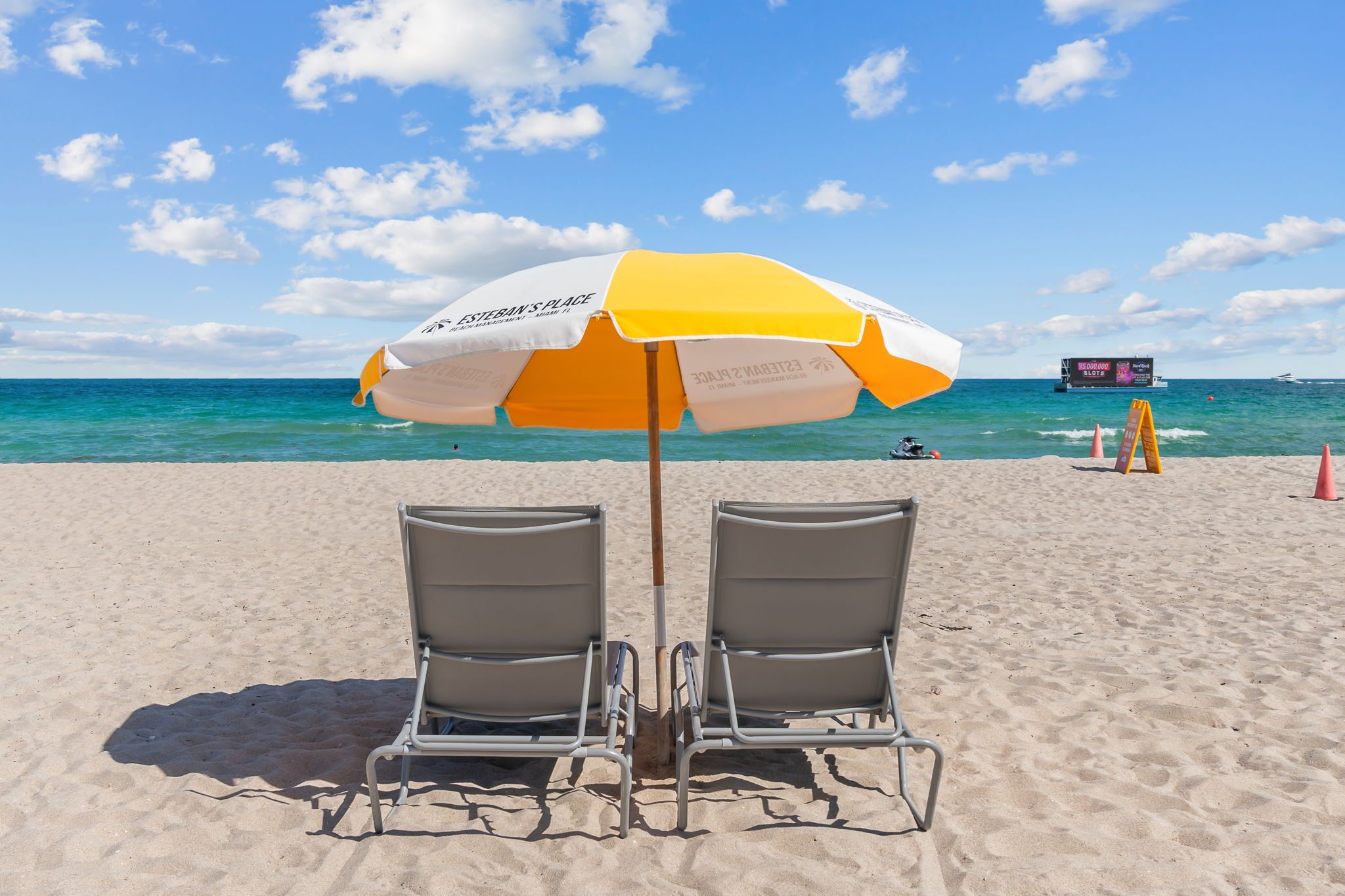 Duas cadeiras de praia sob um guarda-sol amarelo e branco com a marca 'ESTEBAN'S PLACE', em uma praia arenosa com céu limpo e vista para o oceano.