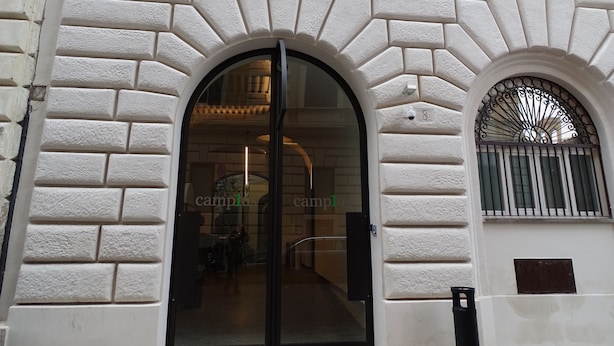 Entrada de edifício com fachada de pedra, porta em arco e janela com grades de metal.