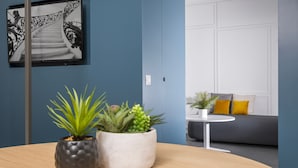 Quarto moderno com parede azul, uma fotografia em preto e branco de uma escadaria emoldurada, uma mesa de madeira redonda com duas plantas em vasos e um sofá cinza com almofadas amarelas.
