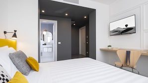 Quarto de hotel moderno com cama, escrivaninha, cadeira, espelho e quadro na parede.