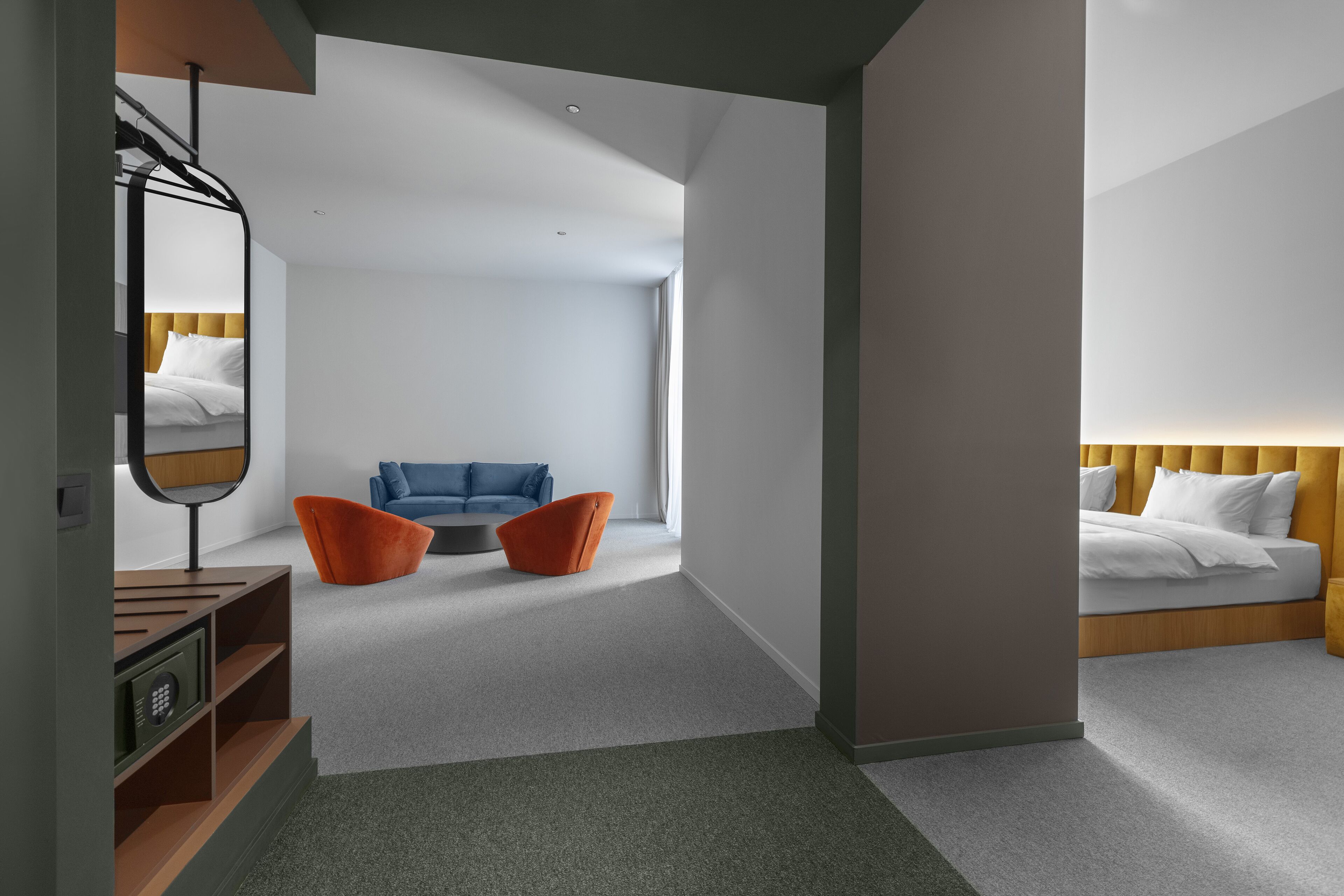 Quarto de hotel moderno com uma cama, área de estar com sofá azul e cadeiras laranja, mesa de cabeceira de madeira e um espelho grande.