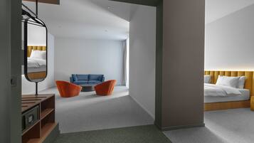 Quarto de hotel moderno com uma cama, área de estar com sofá azul e cadeiras laranja, mesa de cabeceira de madeira e um espelho grande.