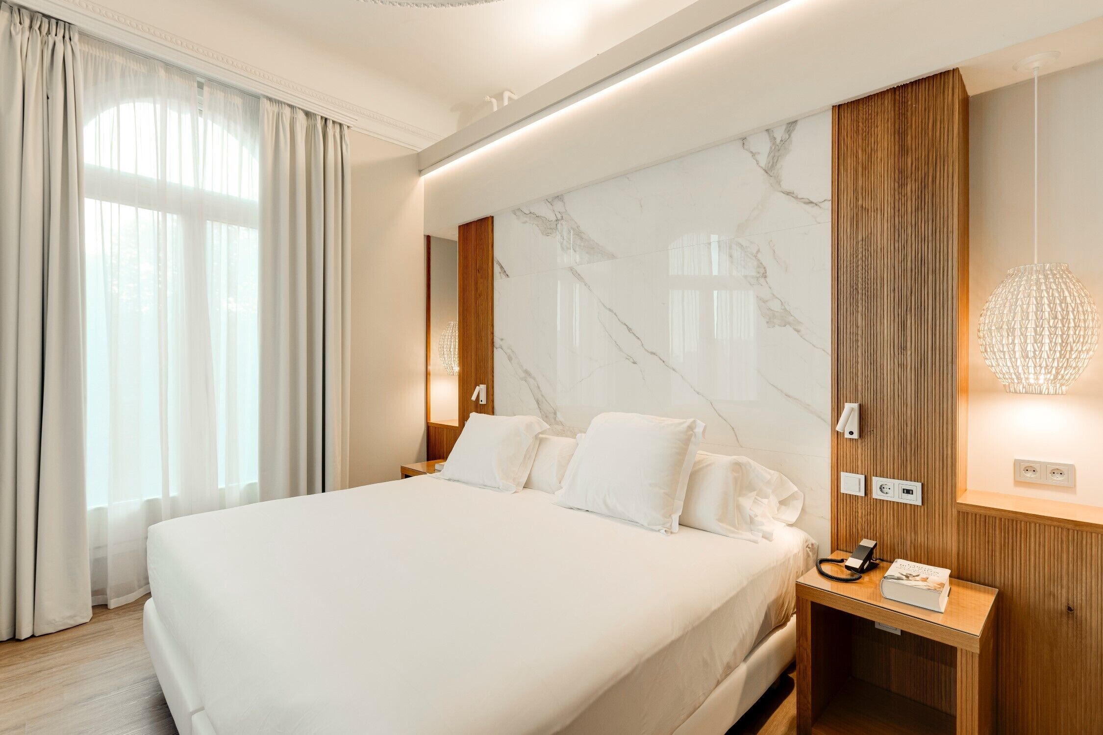 Quarto de hotel moderno com uma cama grande, cabeceira de madeira, parede de mármore e mesa de cabeceira com abajur e telefone.