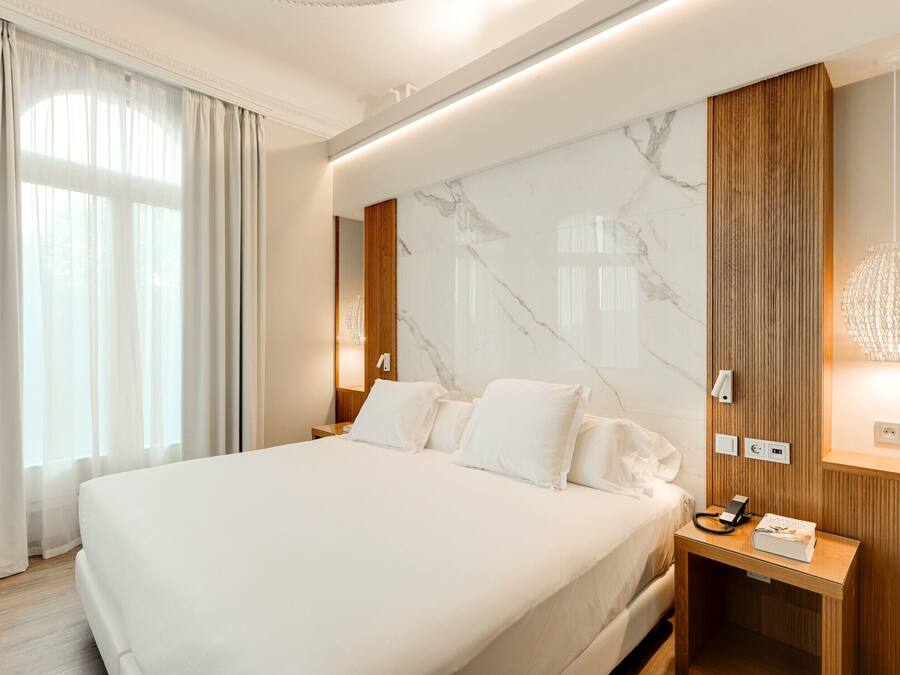 Quarto de hotel moderno com uma cama grande, cabeceira de madeira, parede de mármore e mesa de cabeceira com abajur e telefone.