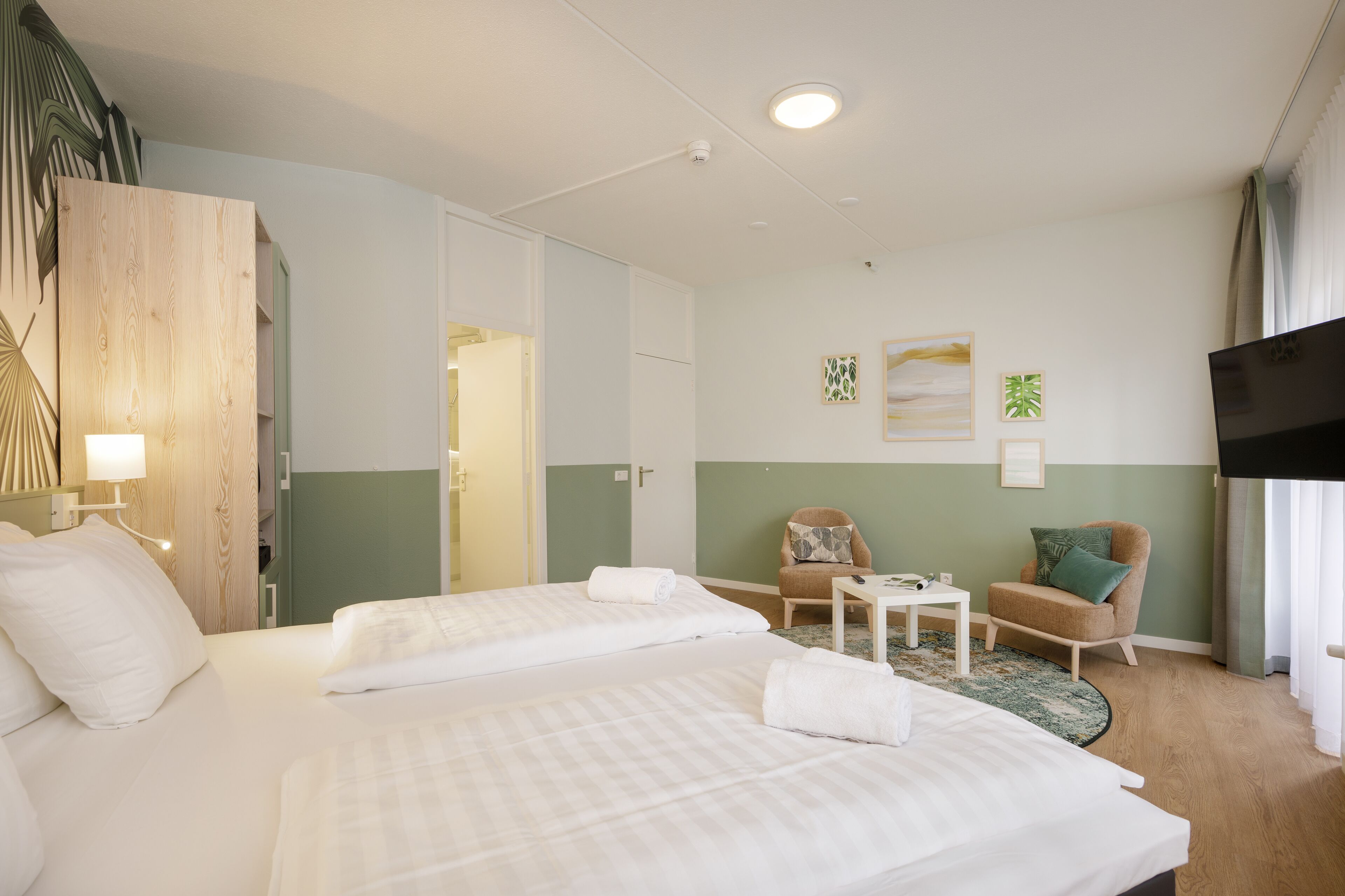 Quarto de hotel com duas camas, uma mesinha, uma cadeira, televisão e obras de arte nas paredes.