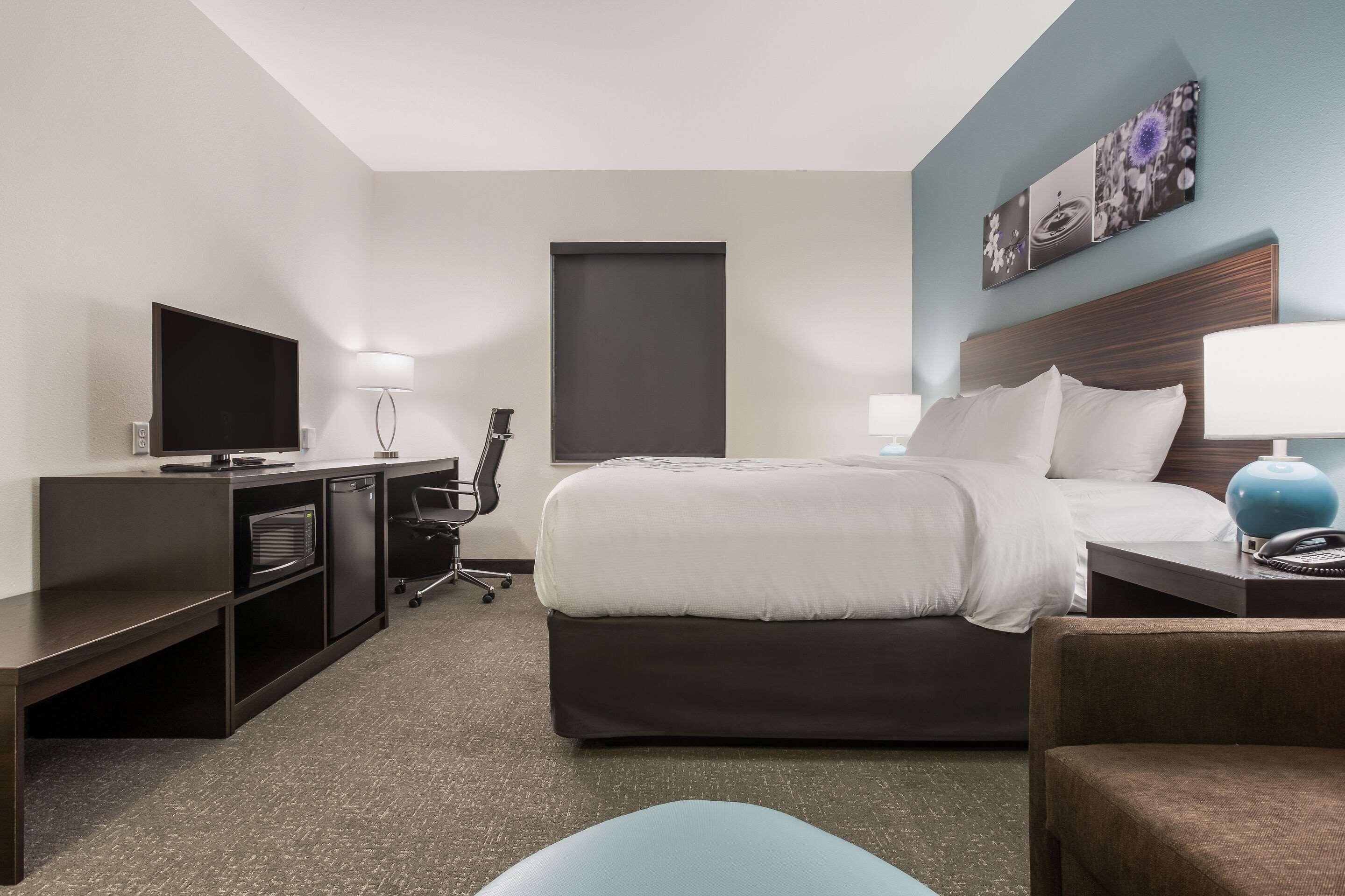 Quarto de hotel com cama, escrivaninha com televisão, cadeira, luminária e uma obra de arte na parede.
