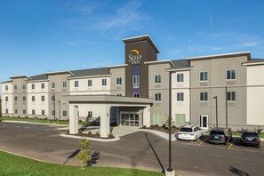Um hotel Sleep Inn com design moderno, apresentando entrada central, vários espaços de estacionamento e céu claro.