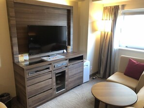 Quarto de hotel com televisão de tela plana sobre um armário de madeira, uma mesa pequena, um sofá e uma janela com cortinas.