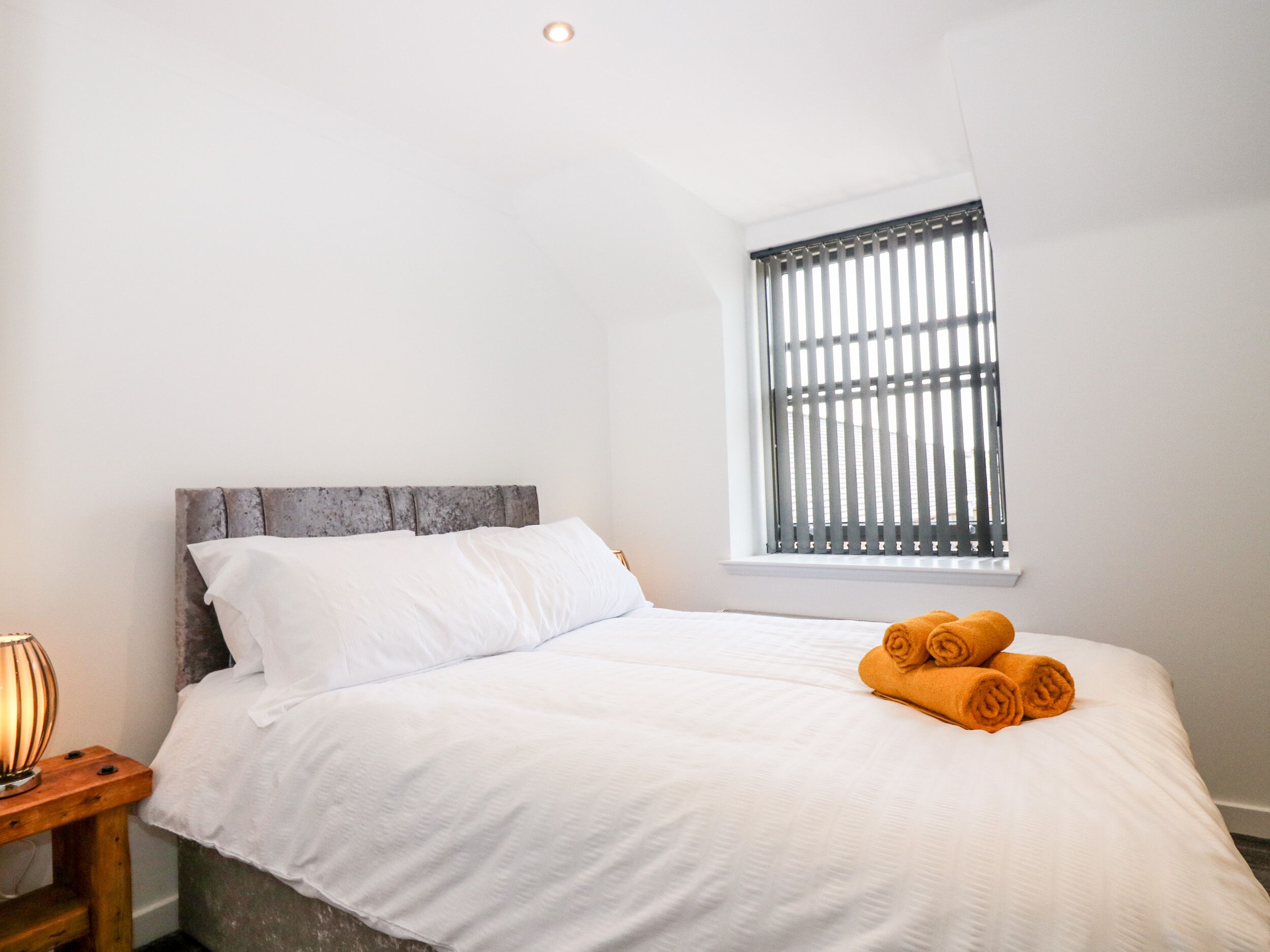 Um quarto com uma cama, uma janela com persianas, um abajur e uma toalha enrolada na cama.