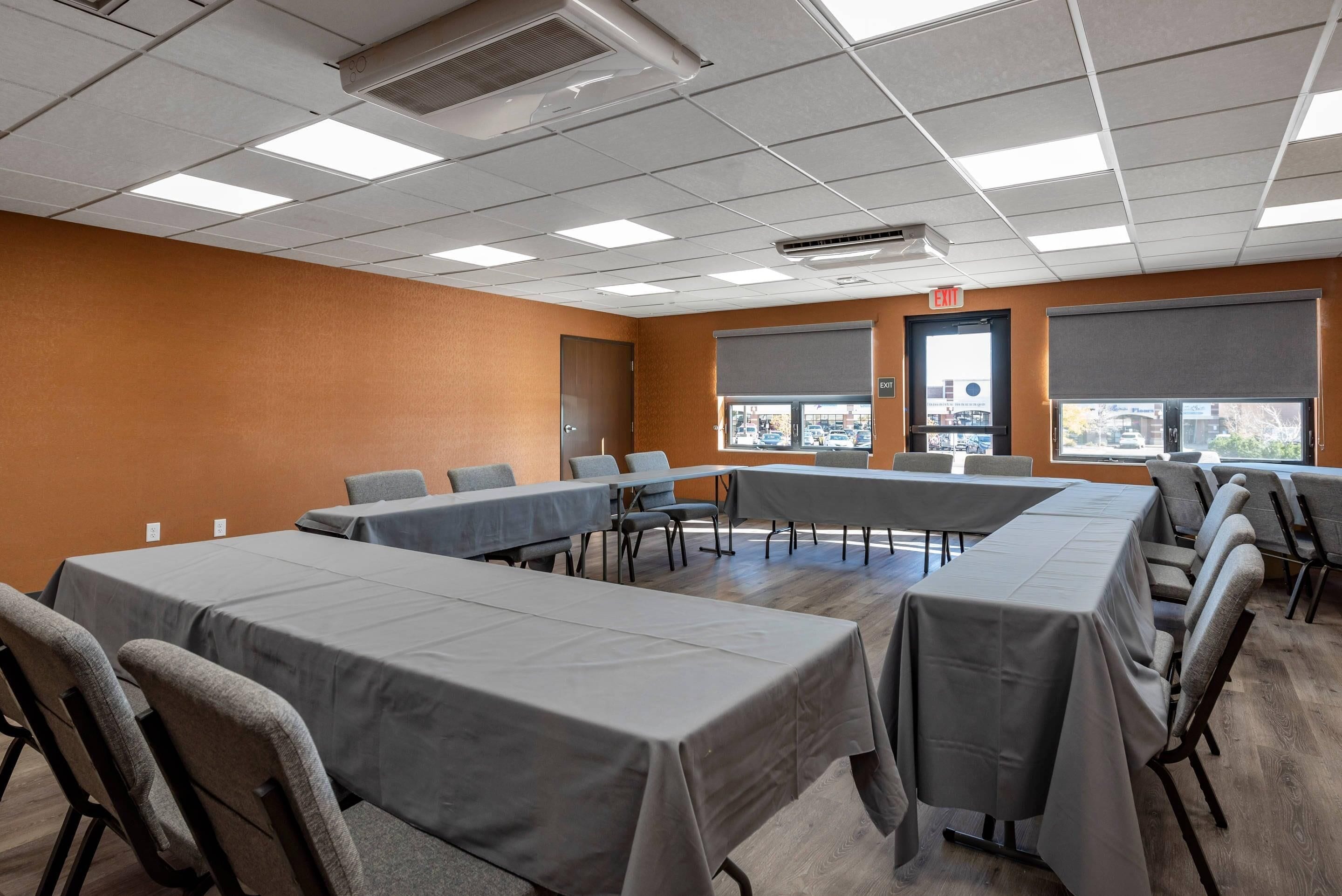 Sala de conferência com mesas e cadeiras, grandes janelas e sinalização de saída.