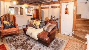 Uma sala de estar aconchegante com um sofá de couro, uma cadeira de madeira e uma mesa de jantar. Há um violino pendurado na parede e uma escada que leva a outro cômodo.