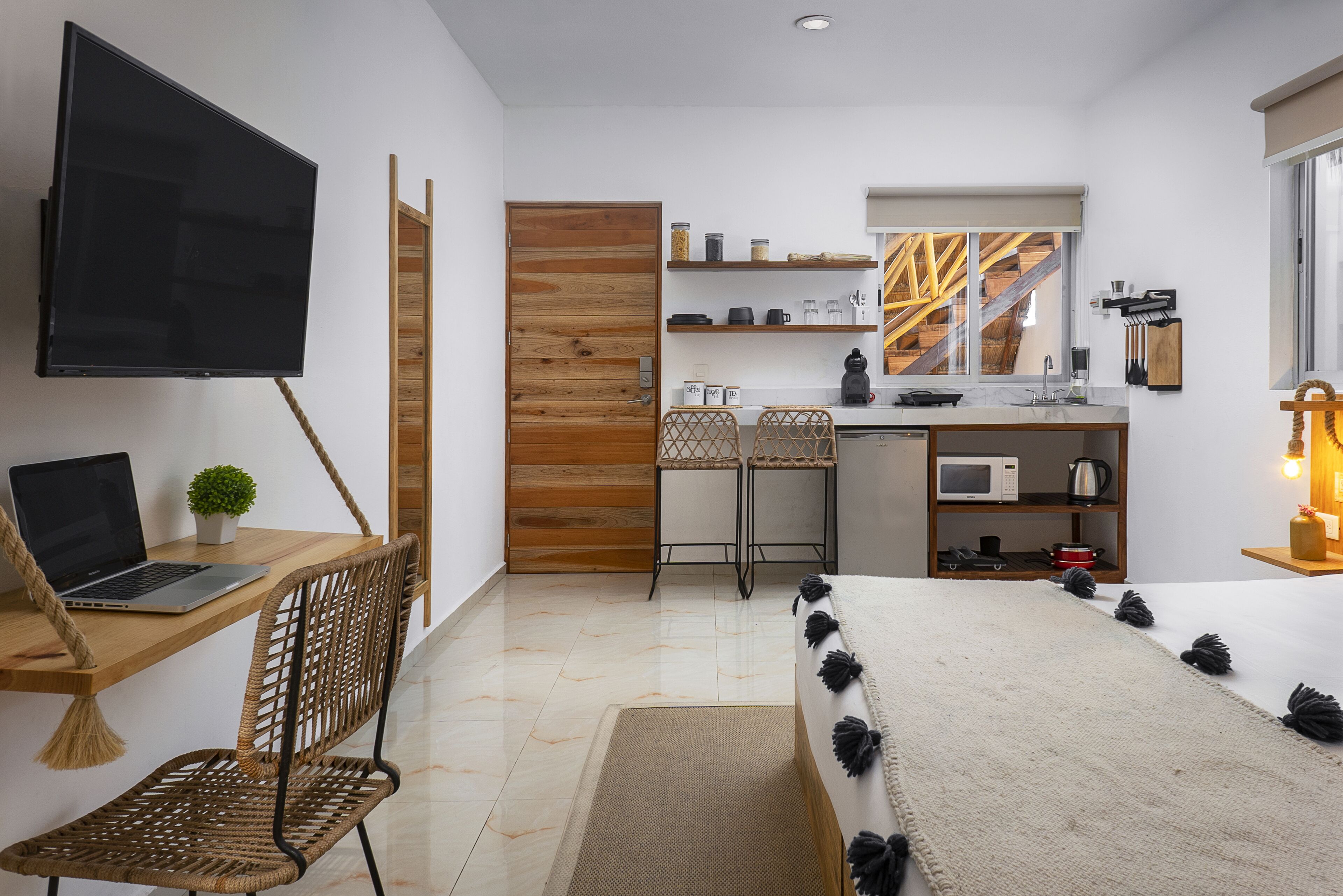Quarto moderno com escrivaninha de madeira, um laptop, uma TV, cama com almofadas decorativas, área de cozinha com micro-ondas e porta de madeira.