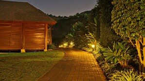 Um edifício de madeira com telhado de tijolos, rodeado por um caminho de jardim bem iluminado e diversas plantas.