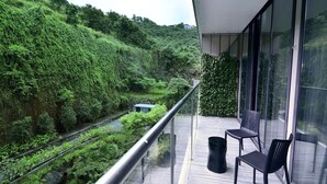 Uma varanda com vista para uma encosta verdejante e uma linha de trem. Há duas cadeiras pretas e uma lixeira preta na varanda.