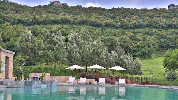 Uma piscina com espreguiçadeiras e guarda-sóis, cercada por vegetação exuberante e um campo de golfe ao fundo.