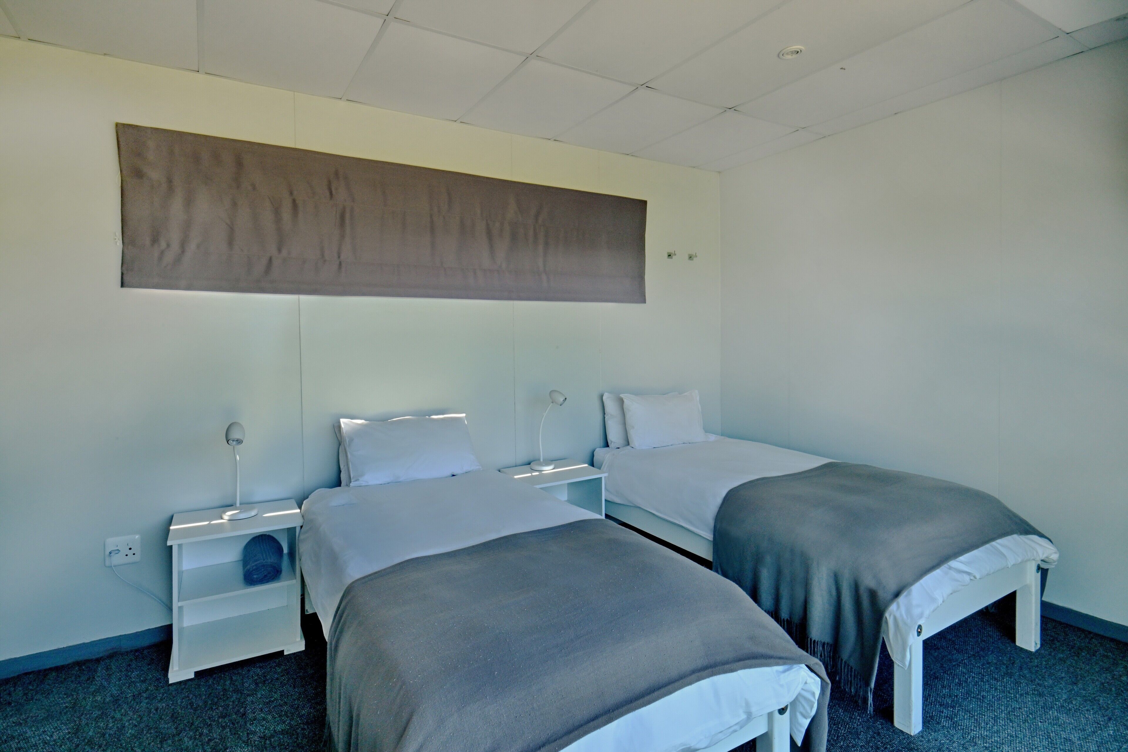 Quarto de hotel com duas camas de solteiro, uma mesa de cabeceira com abajur e uma janela grande com persiana bege.