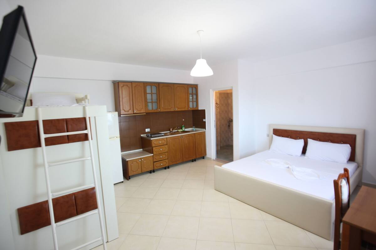 Quarto com cama, pequeno espaço de cozinha e televisão sobre suporte.