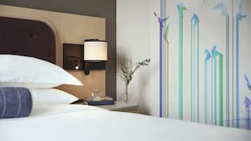 Quarto de hotel com cama, mesa de cabeceira, abajur e parede com listras verticais e ilustrações de pássaros.
