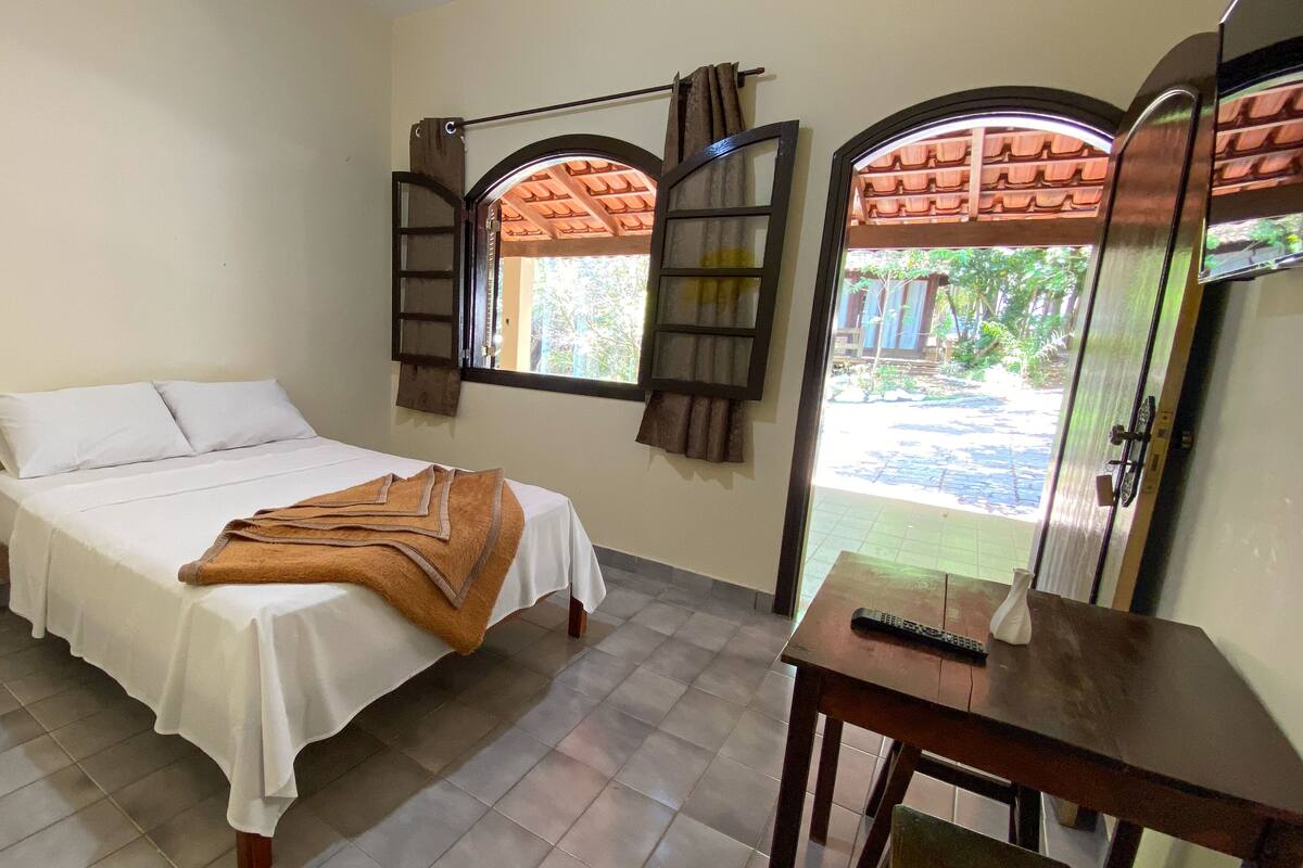 Quarto com cama, escrivaninha e cadeira. Duas janelas com as persianas abertas.