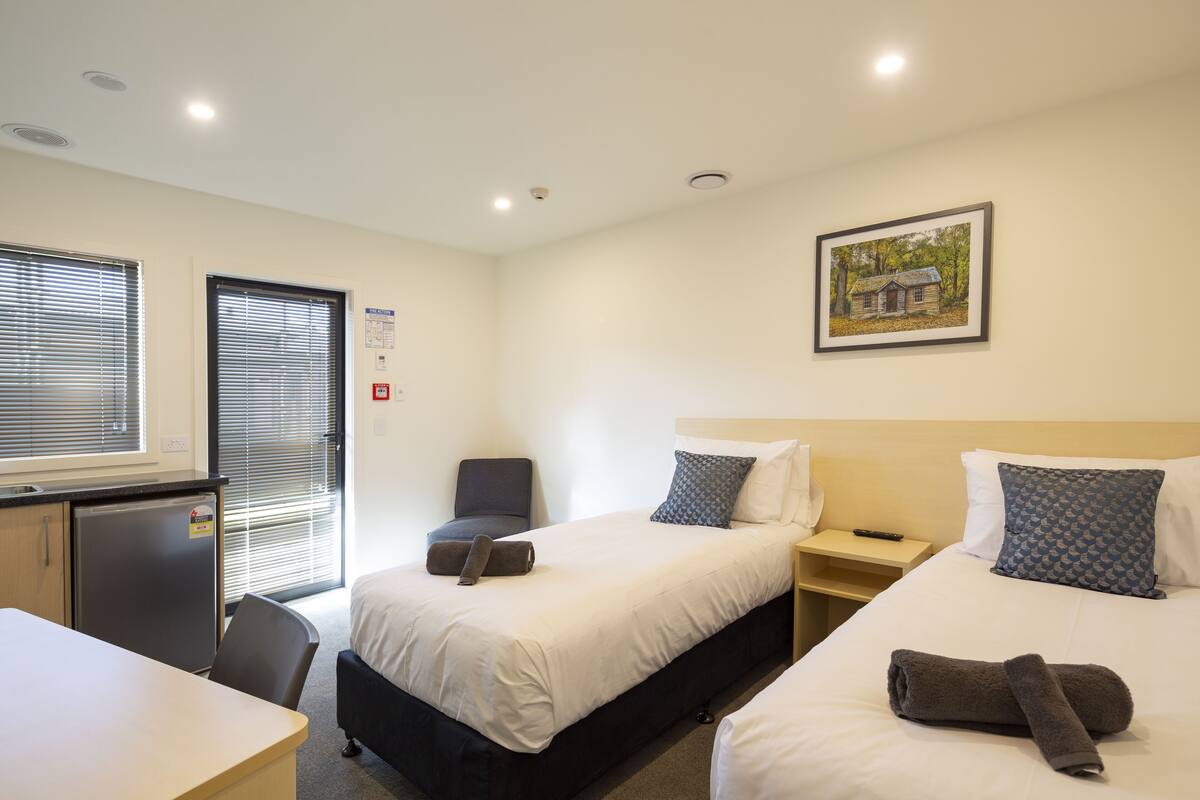 Quarto de hotel com duas camas, uma escrivaninha, um frigorífico e uma janela com persianas.