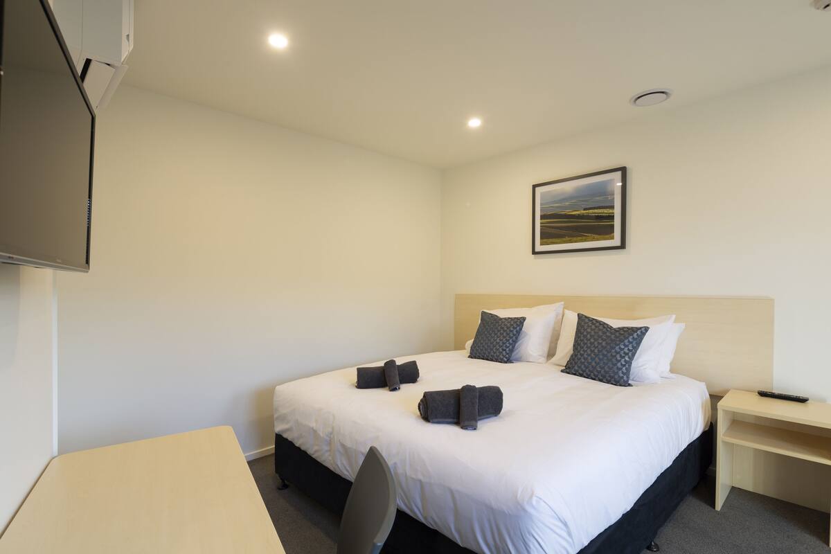 Quarto de hotel com cama, escrivaninha, cadeira, mesa de cabeceira e televisão na parede.