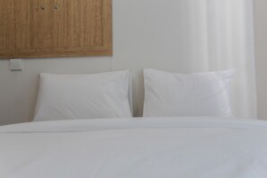Uma cama com roupa de cama branca e duas almofadas, um armário de madeira em cima e uma parede branca.